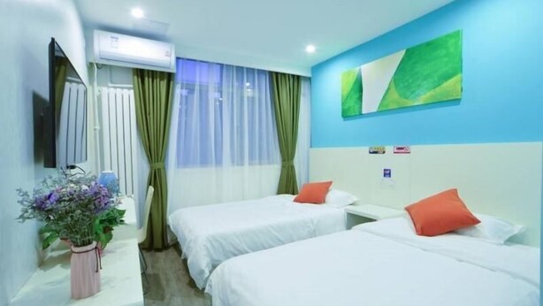 Quarto de hotel com duas camas, uma televisão de tela plana, uma janela com cortinas translúcidas, uma planta verde e um quadro abstrato azul na parede.