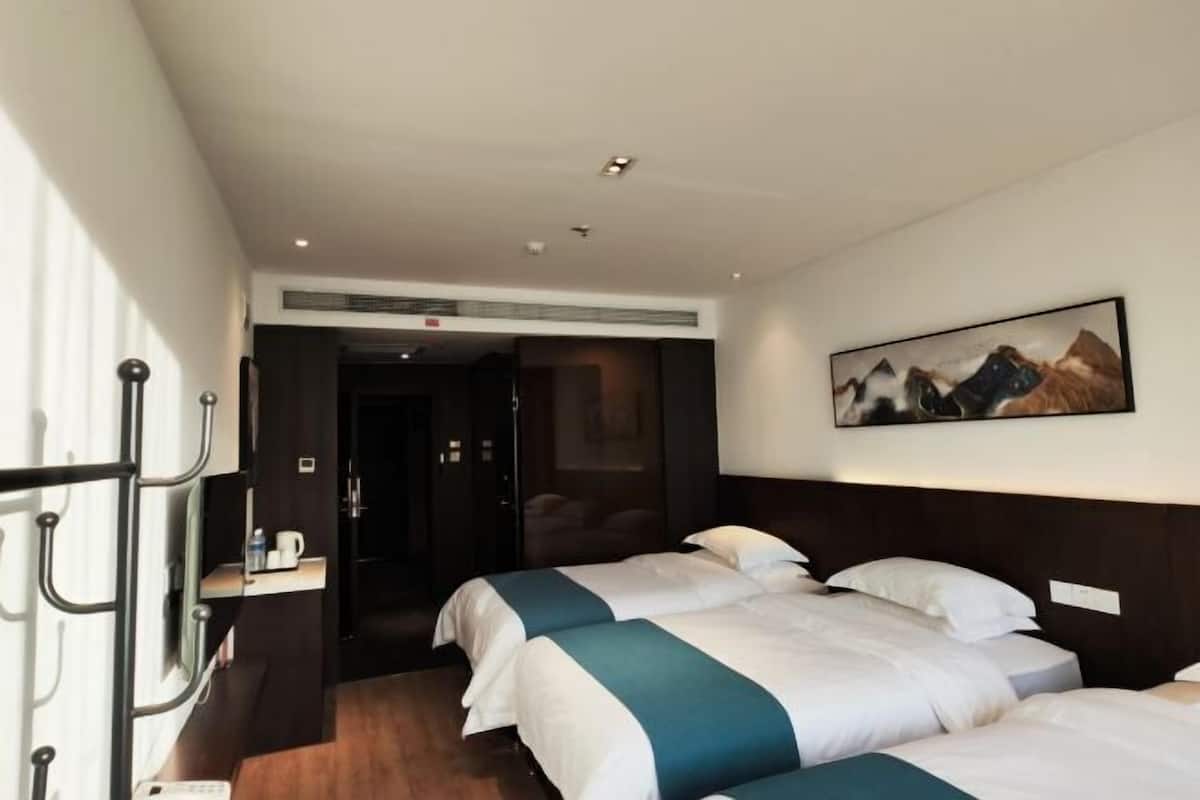 Quarto de hotel com duas camas, uma pintura de montanhas na parede, uma pequena prateleira com uma garrafa de água e uma porta que dá para outro quarto.