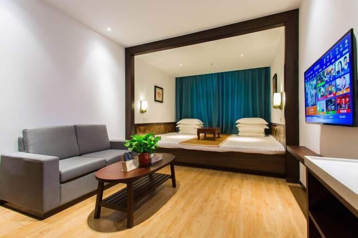 Quarto de hotel com sofá, uma pequena mesa com uma planta, cama com almofadas, um espelho grande e TV na parede.