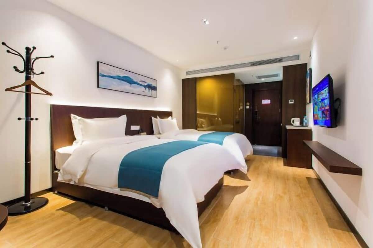 Quarto de hotel com duas camas, um cabideiro, um quadro, uma televisão e piso de madeira.