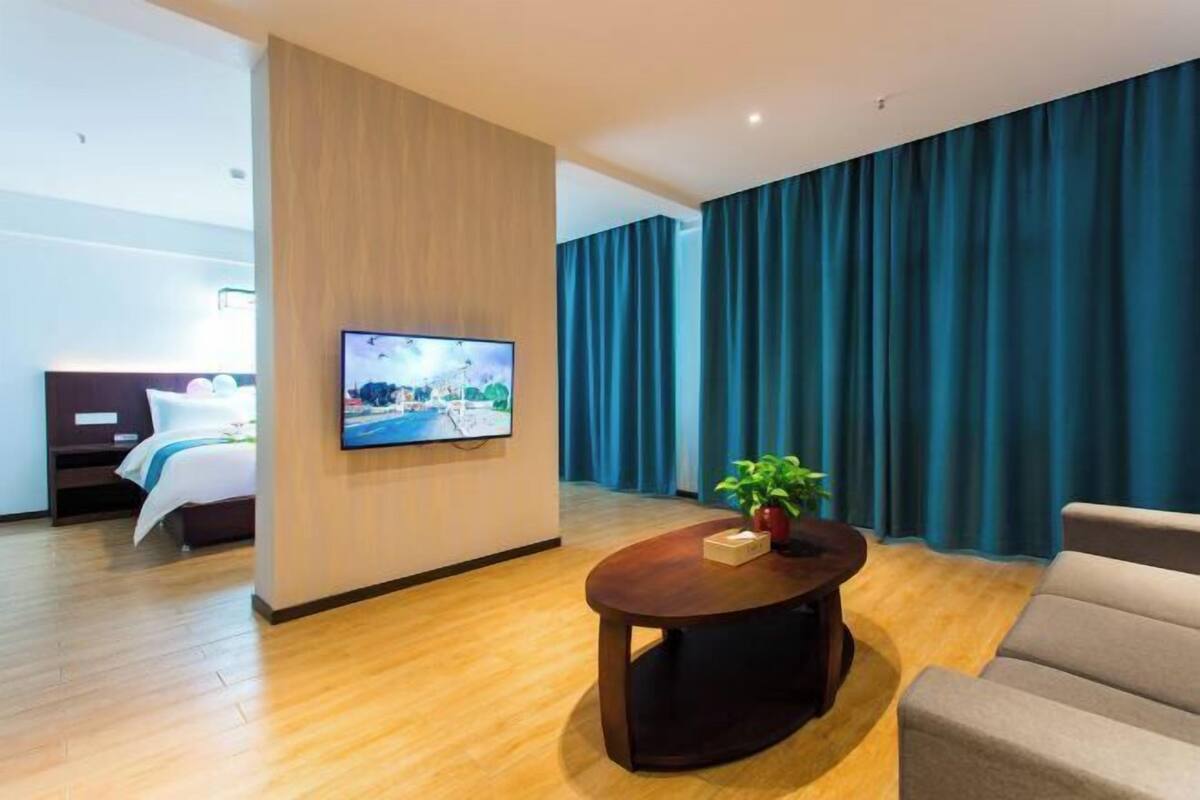Quarto de hotel moderno com piso de madeira, cama com roupa de cama branca, TV de tela plana na parede, mesa de centro redonda com uma planta e uma grande janela com cortinas azuis.