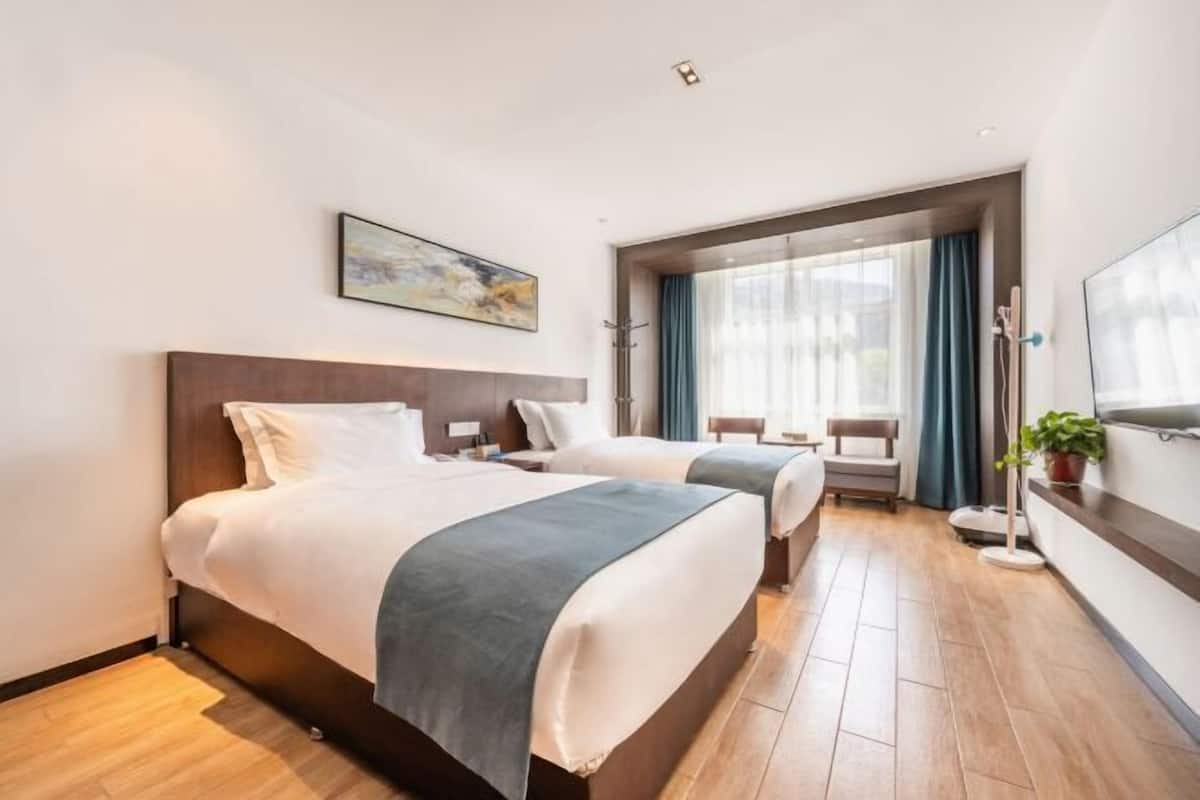 Quarto de hotel com duas camas, uma janela grande com cortinas, uma televisão de tela plana e um quadro na parede.