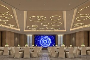 Salão de festas com mesas redondas, palco central e teto com luminárias circulares.