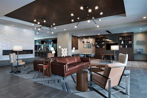 Um lobby de hotel moderno com lareira, sofás de couro, área de refeições e cozinha ao fundo.