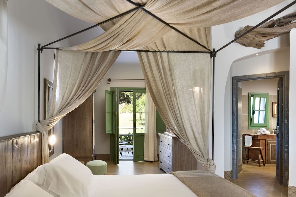 Quarto com cama de dossel, cômoda e vista do jardim através de uma porta aberta.