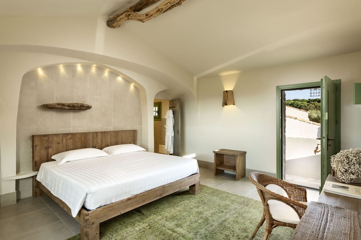 Quarto com cama de madeira, tapete verde, cadeira de vime e vista para o exterior.