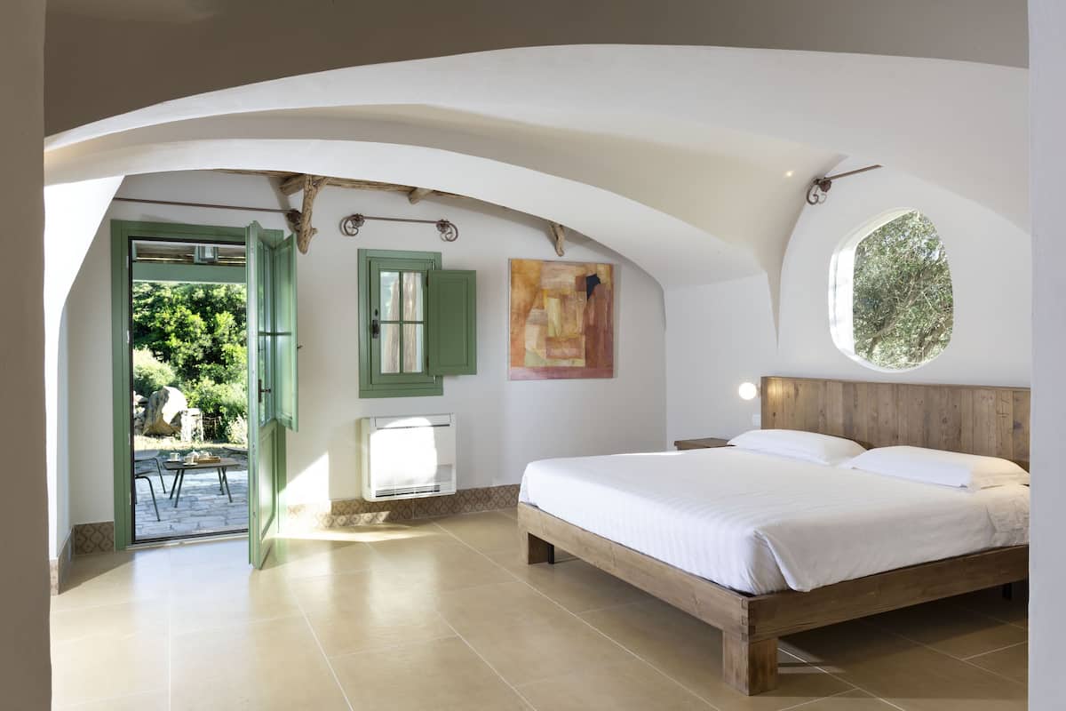 Quarto com cama de madeira, janela com moldura verde e vista para o verde lá fora.