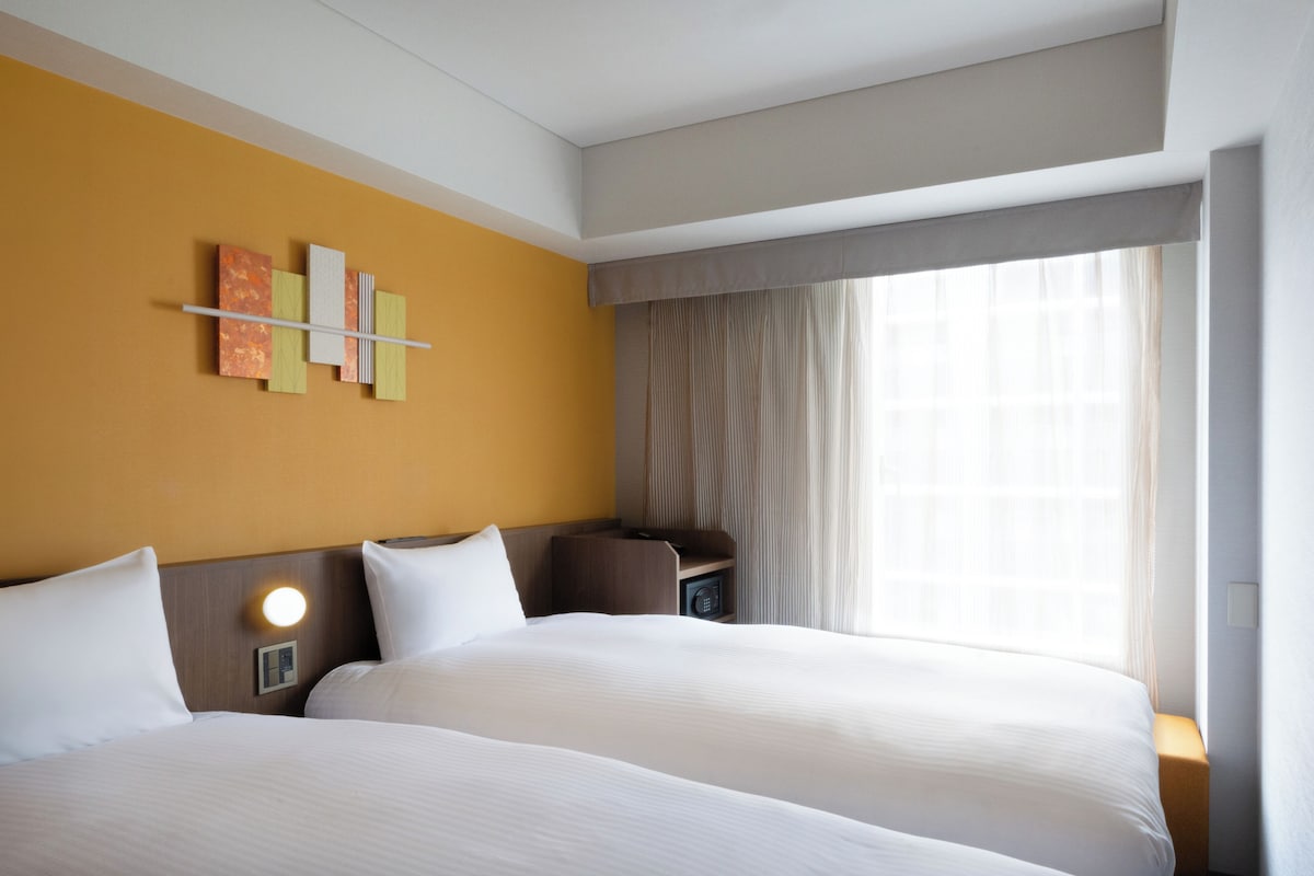 Quarto de hotel com duas camas, mesa de cabeceira, luminária na parede e janela com cortinas.