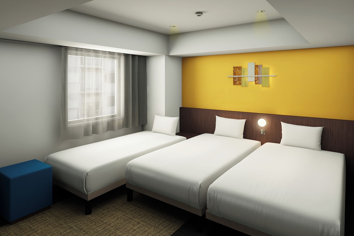 Quarto de hotel com duas camas de solteiro, um banquinho pequeno azul, uma janela grande com cortinas translúcidas e uma parede amarela com obras de arte.
