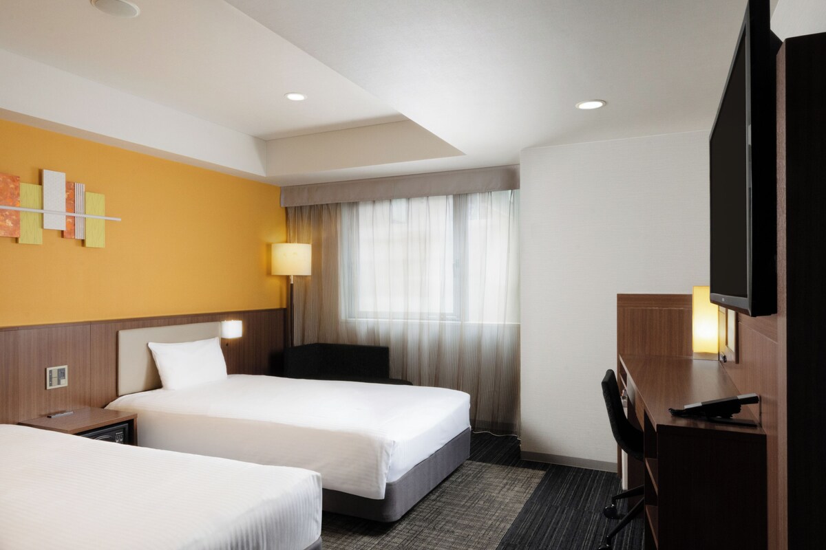 Quarto de hotel com duas camas, uma escrivaninha, uma televisão e uma janela com cortinas.