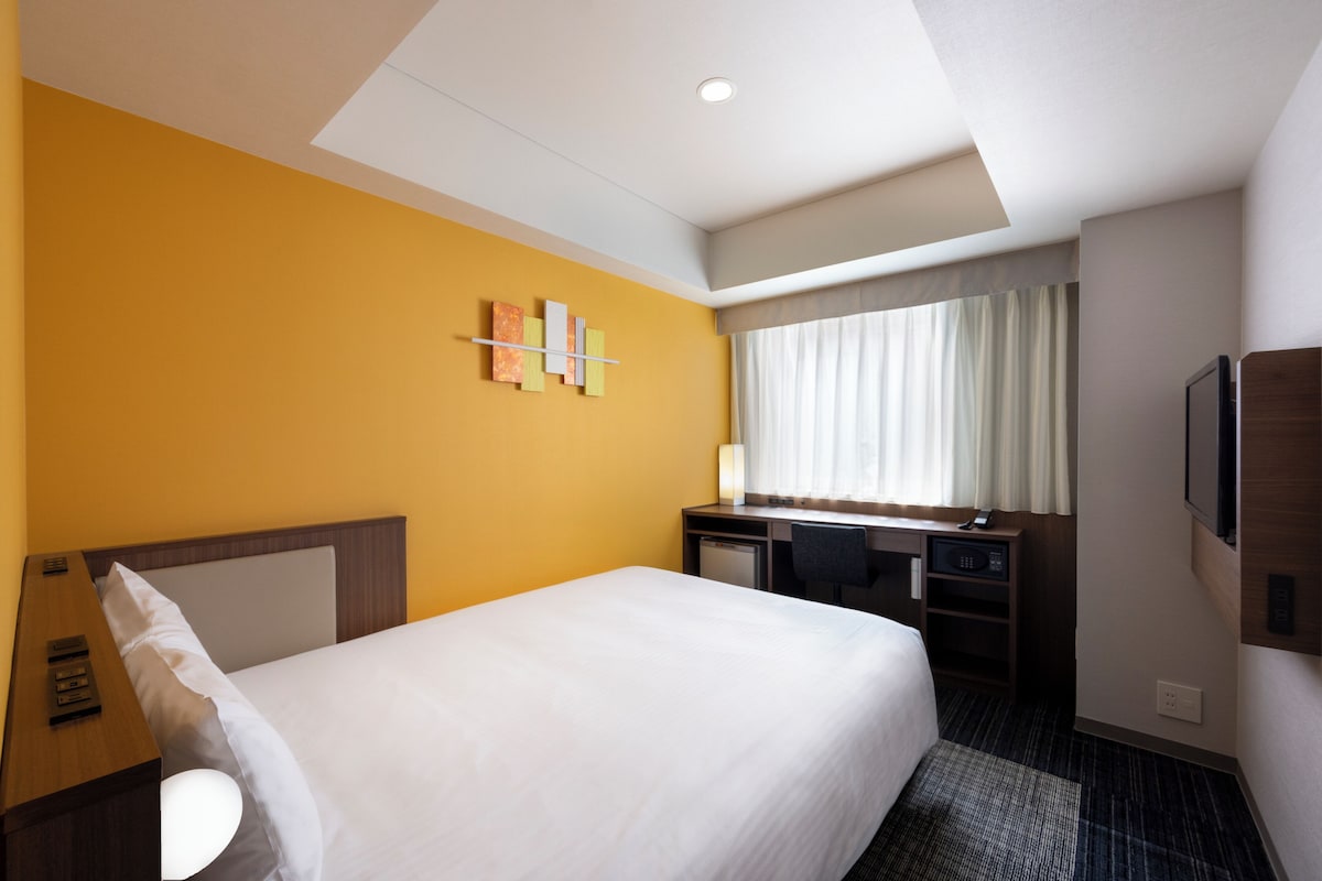 Quarto de hotel com cama, escrivaninha e janela com cortinas.