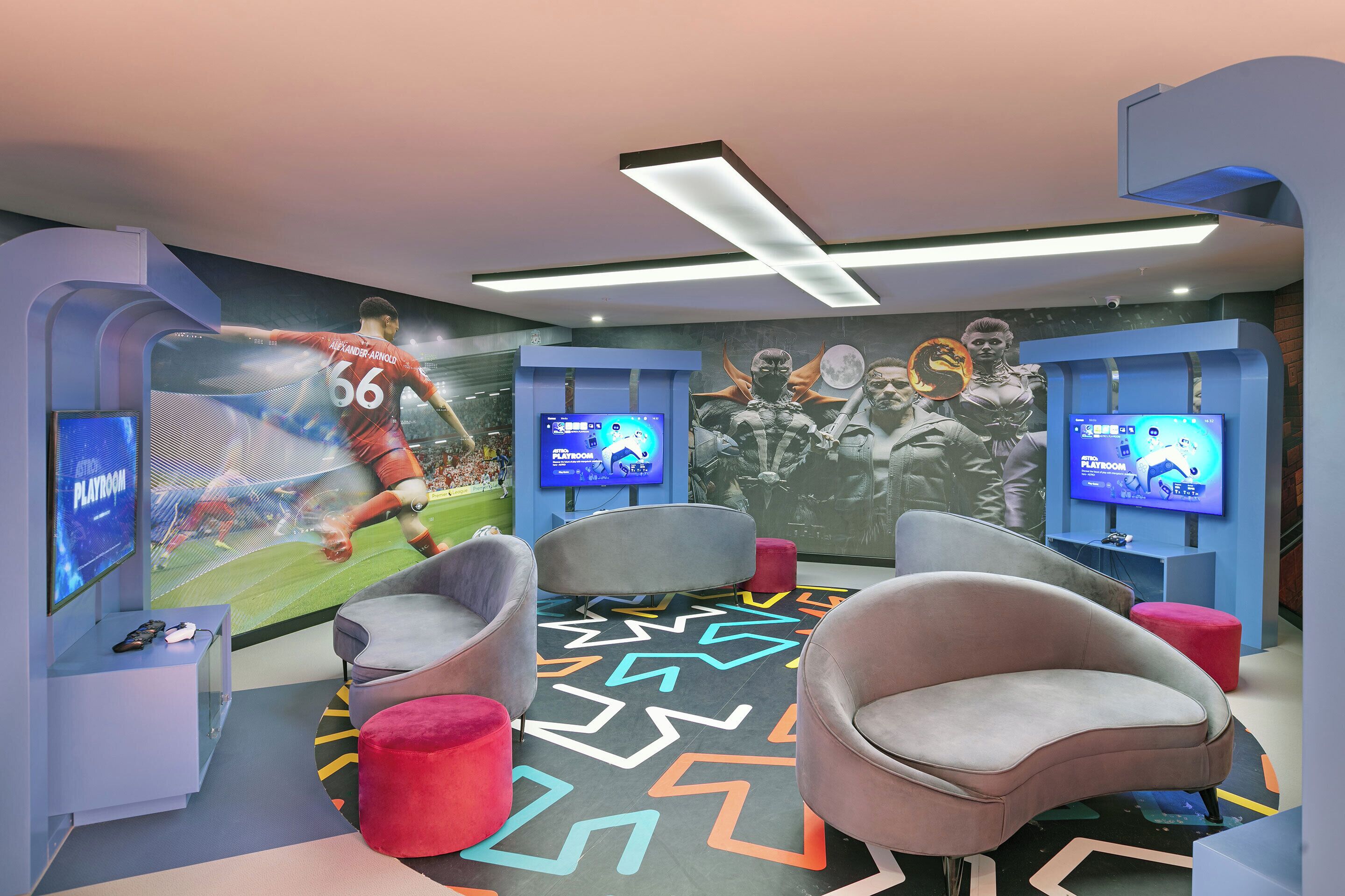 Uma sala de jogos moderna com várias telas, assentos confortáveis e um mural vibrante na parede.