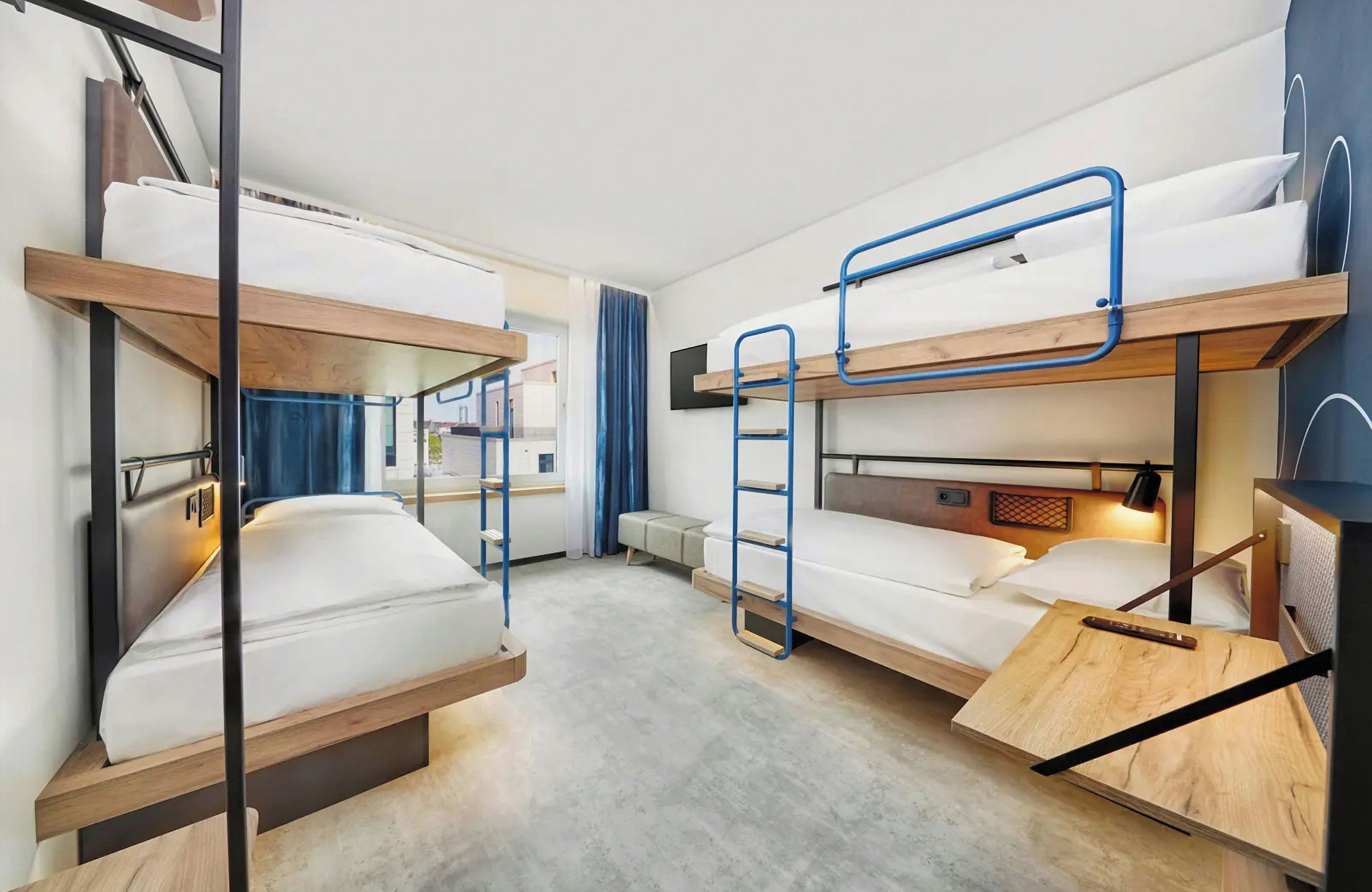 Quarto de hotel pequeno com beliches, uma escrivaninha e uma janela com cortinas.