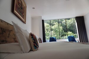Quarto de hotel com uma cama grande, janela com vista para árvores, uma cadeira azul e um quadro na parede.