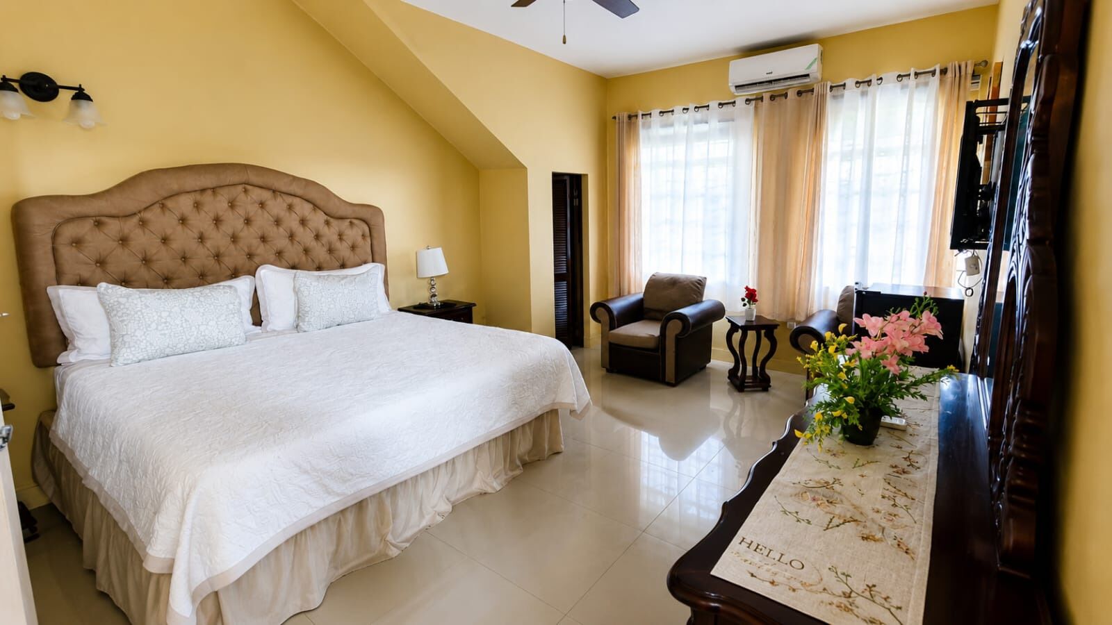 Quarto de hotel com uma cama grande, uma cadeira, uma mesinha com flores e uma escrivaninha com um aviso.