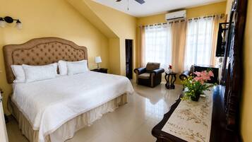 Quarto de hotel com uma cama grande, uma cadeira, uma mesinha com flores e uma escrivaninha com um aviso.