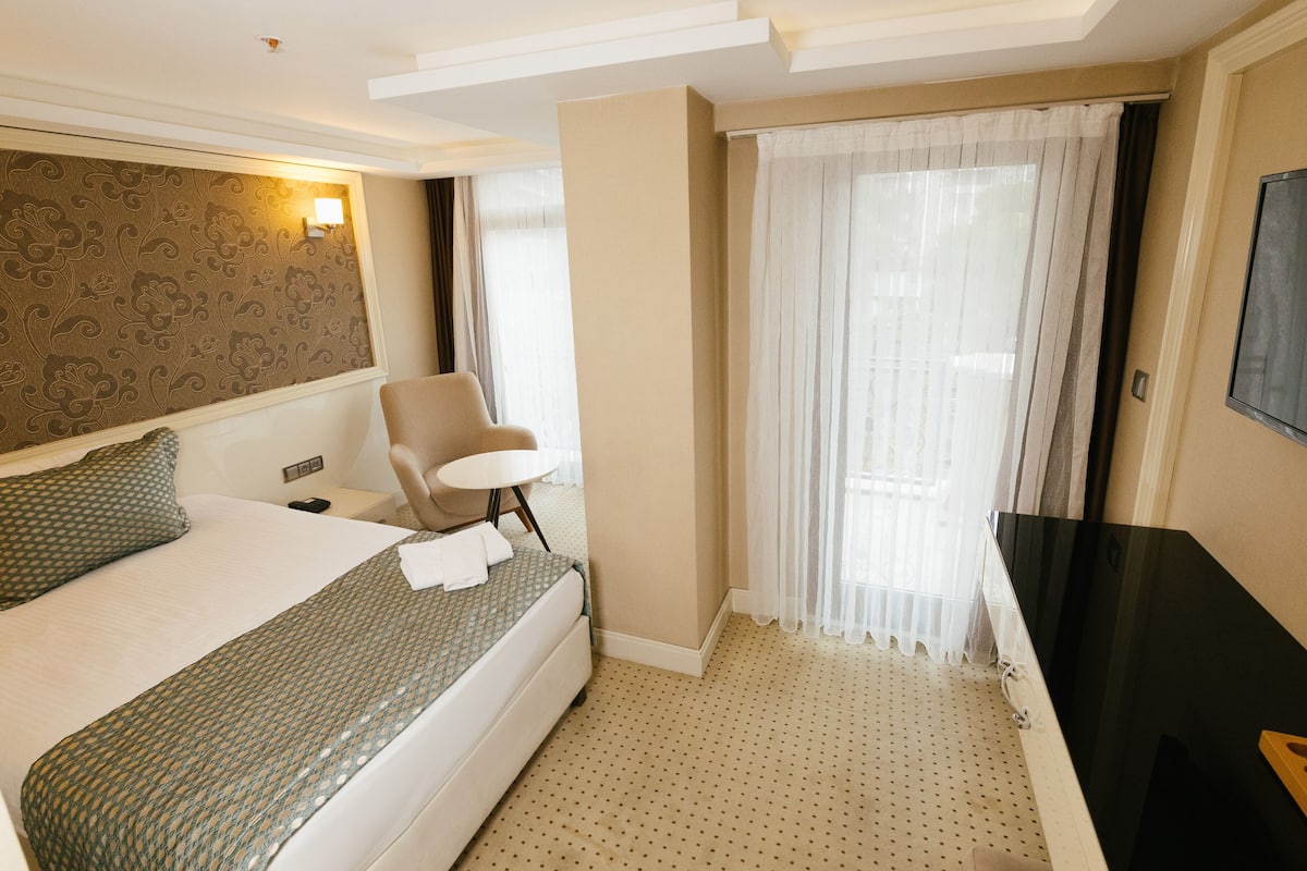 Quarto de hotel com cama, poltrona, mesinha, televisão e janela com cortinas.