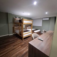 Quarto com beliche, escrivaninha, cadeira, piso de madeira e quadro branco na parede.