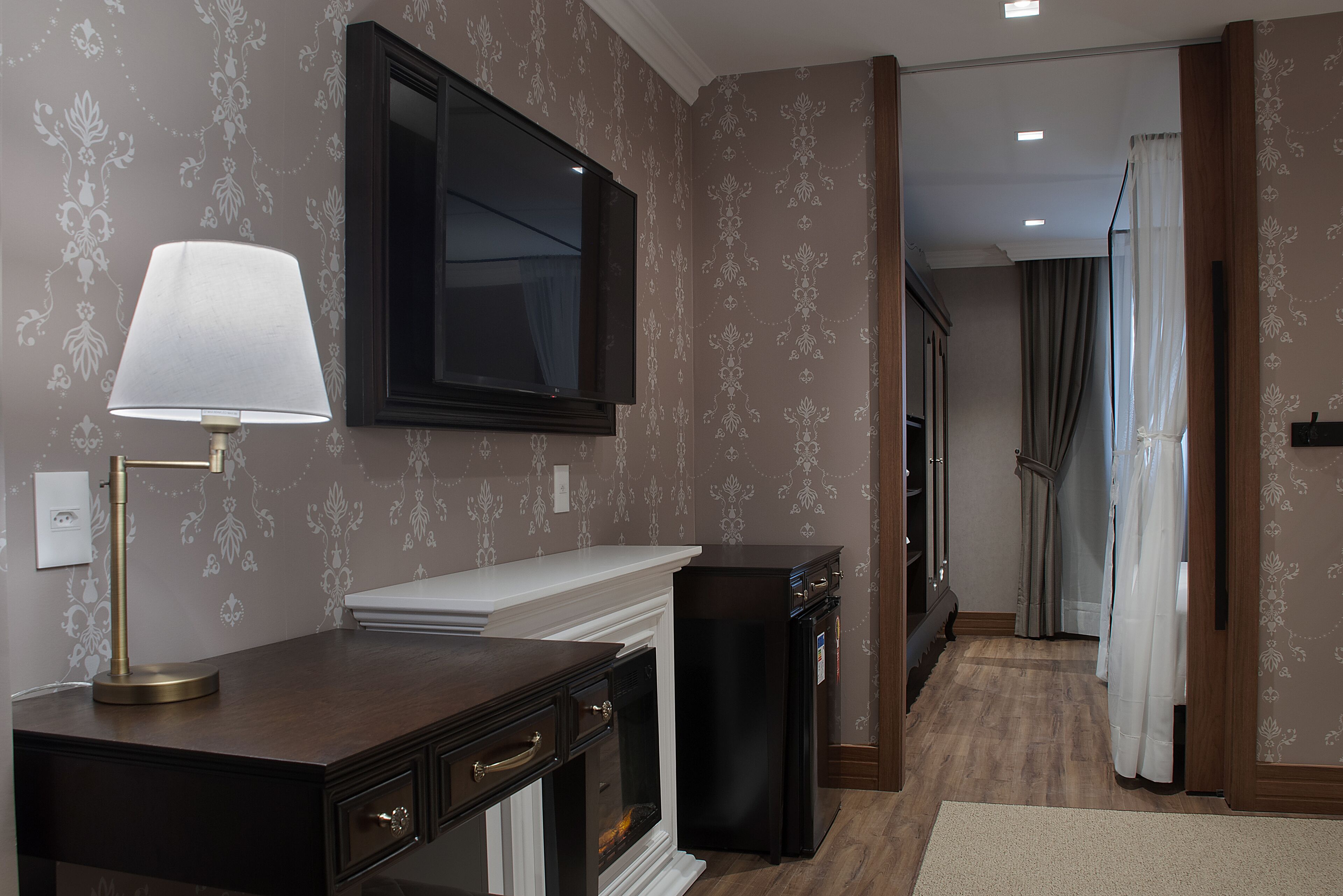 Quarto de hotel com televisão de tela plana, escrivaninha com gavetas, luminária, geladeira e banheiro visível por uma porta aberta.