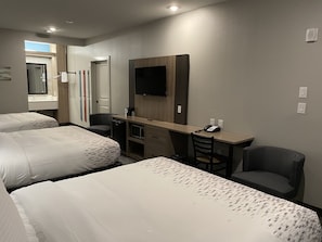 Quarto de hotel com duas camas, uma escrivaninha, uma cadeira, televisão e uma pequena cozinha.