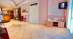 Um saguão de hotel com balcão de recepção, área de refeições, televisão na parede e piso revestido de azulejos.