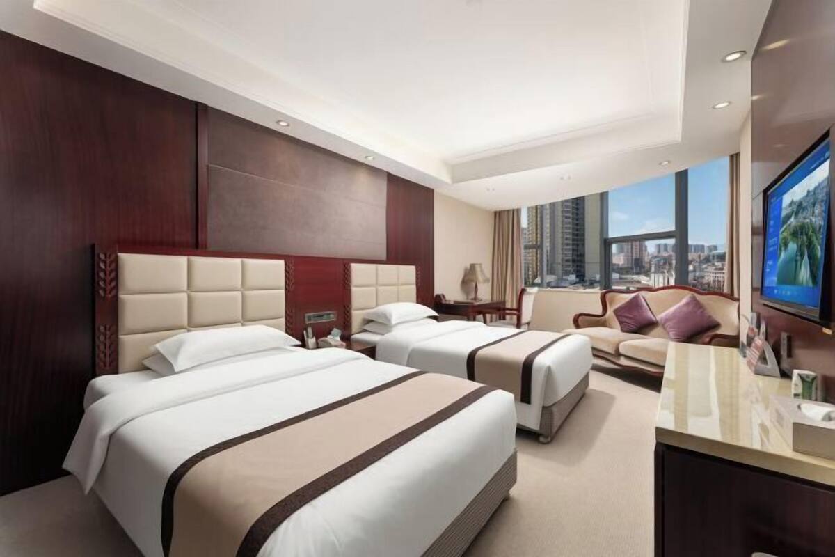 Quarto de hotel com duas camas, uma mesa, uma cadeira e uma janela grande com vista para a cidade.
