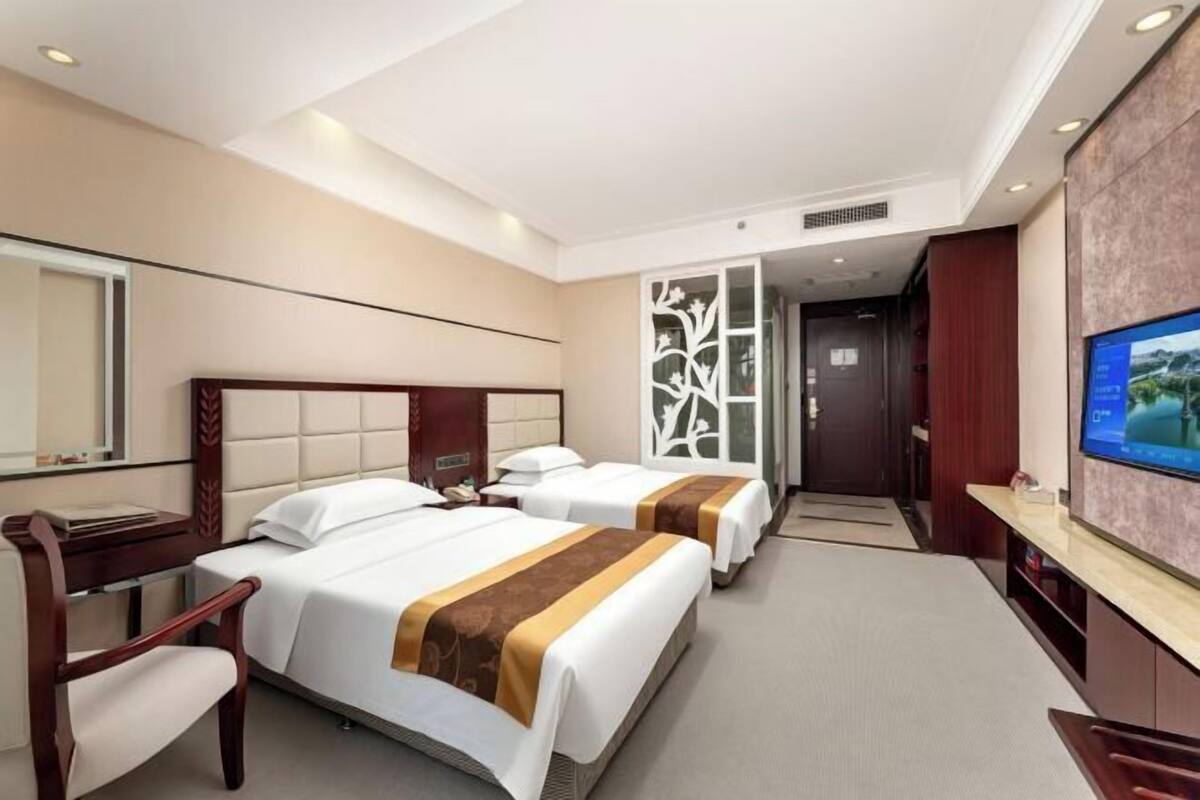 Quarto de hotel com duas camas, uma escrivaninha, uma cadeira, uma TV e um painel decorativo na parede.