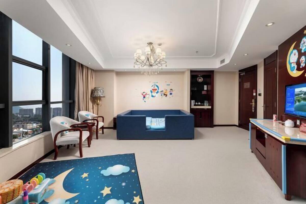 Quarto de hotel com uma janela grande, um sofá azul, uma escrivaninha com um computador e uma televisão.