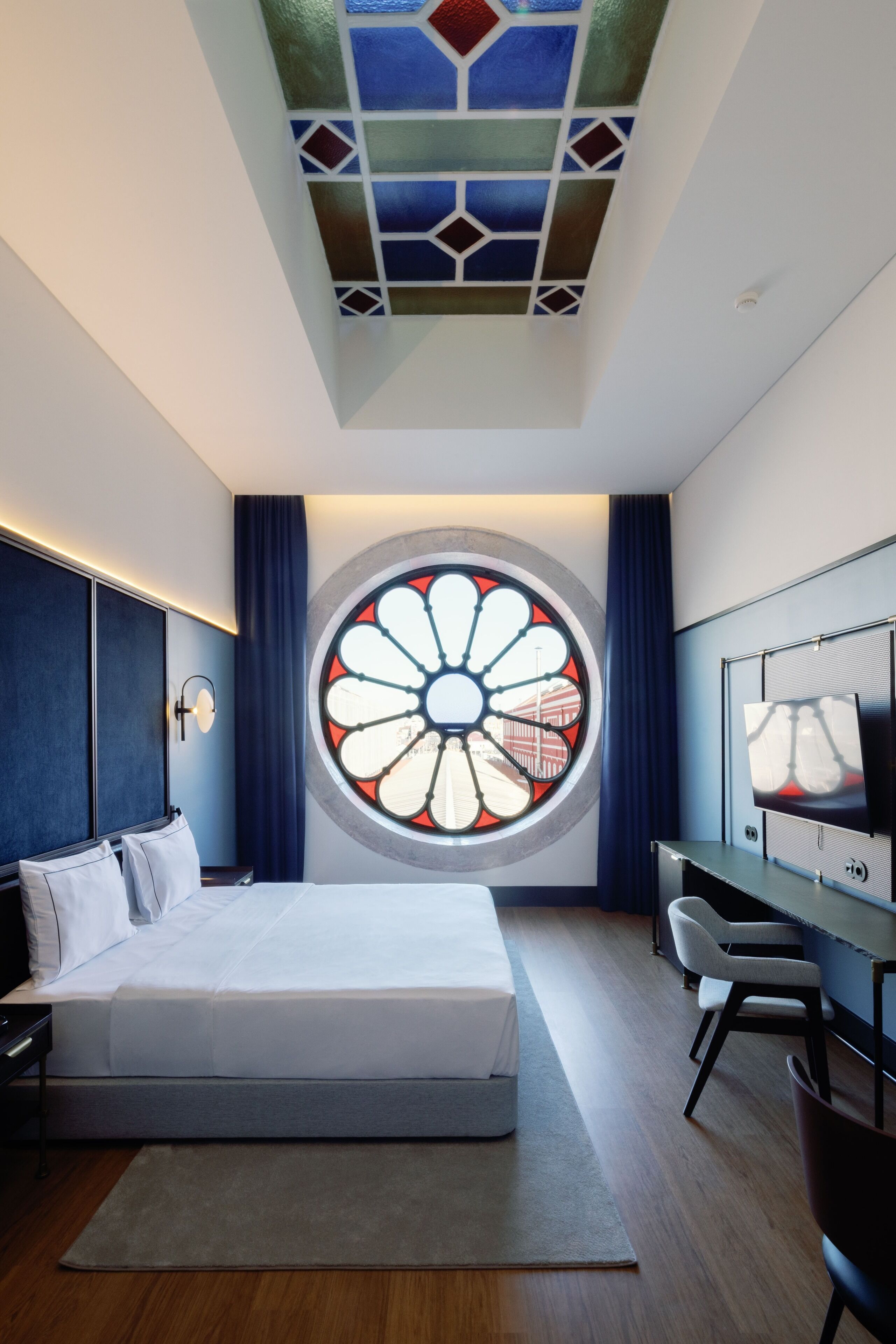 Quarto de hotel moderno com uma cama grande, uma escrivaninha, uma televisão e um vitral exclusivo sobre a cama.