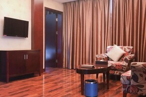 Quarto de hotel com piso de madeira, televisão na parede, sofá com estampa floral, mesa pequena e lixeira azul.