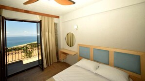 Quarto de hotel com cama, mesa de cabeceira, espelho redondo, ventilador de teto e varanda com vista para o mar.