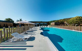 Piscina com espreguiçadeiras e guarda-sóis, cercada por cercas de madeira, e um edifício ao fundo.