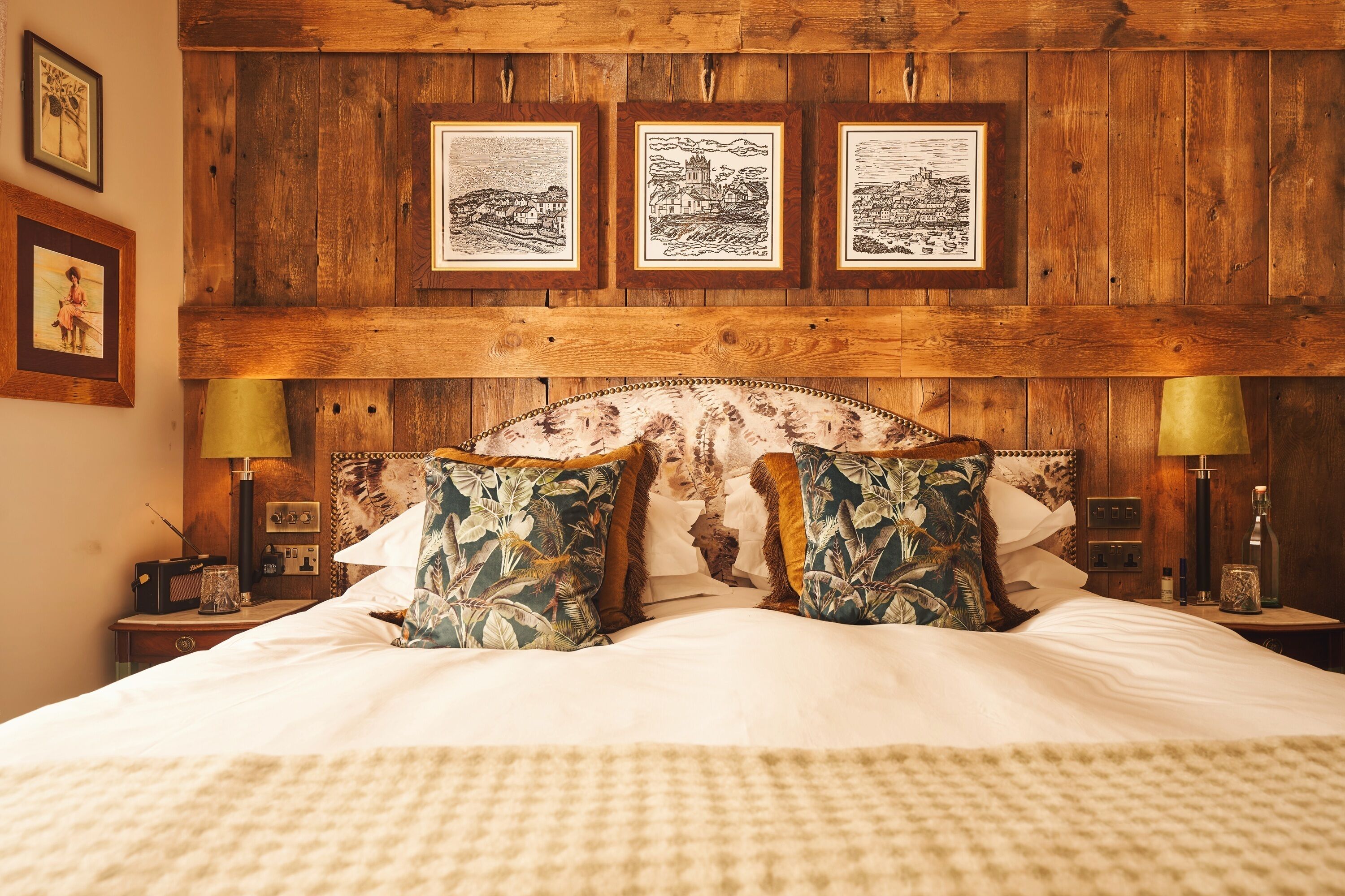 Quarto com cabeceira de madeira, cama com roupa de cama branca, duas mesas de cabeceira com abajures e três quadros na parede.