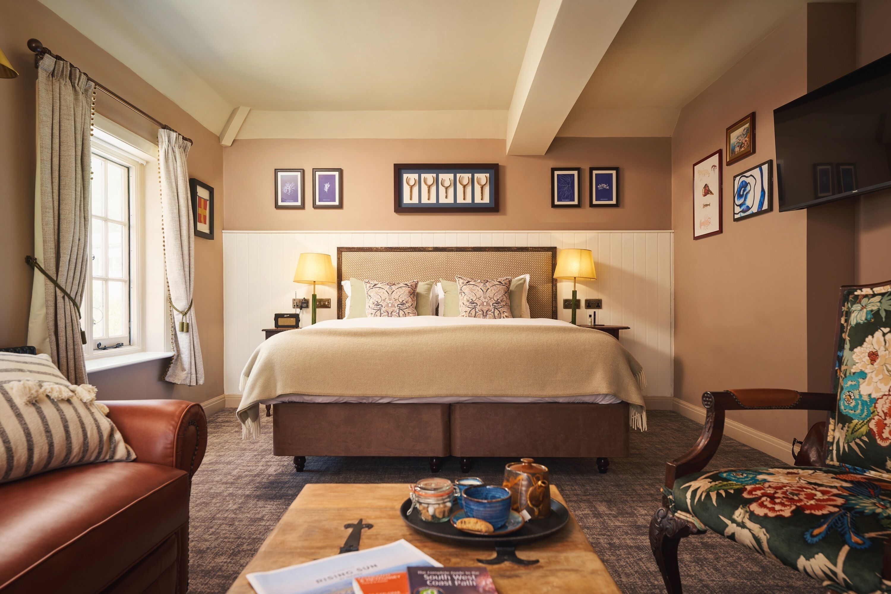Quarto de hotel com uma cama grande, duas luminárias de cabeceira, uma mesa de centro de madeira com uma bandeja de itens de café da manhã, uma poltrona de couro e uma poltrona florida.
