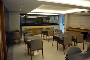 Uma sala de jantar moderna com várias mesas e cadeiras, uma cozinha ao fundo e um aparelho de ar condicionado na parede.
