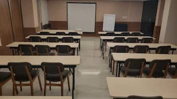 Uma sala de conferências com fileiras de mesas e cadeiras, uma tela de projetor e um quadro branco.
