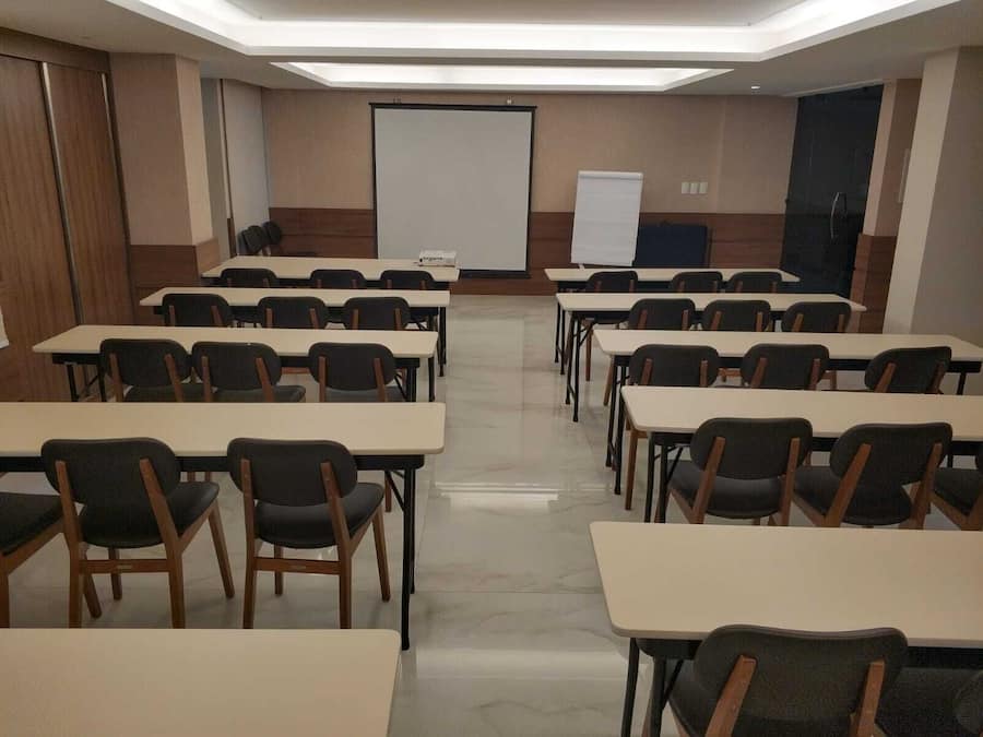Uma sala de conferências com fileiras de mesas e cadeiras, uma tela de projetor e um quadro branco.