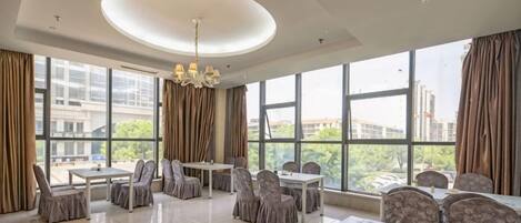 Um espaço de refeições de hotel com uma mesa central, várias cadeiras e grandes janelas com vista para a cidade.