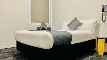 Quarto de hotel com cama, janela, telefone e uma pequena mesa de cabeceira de madeira.