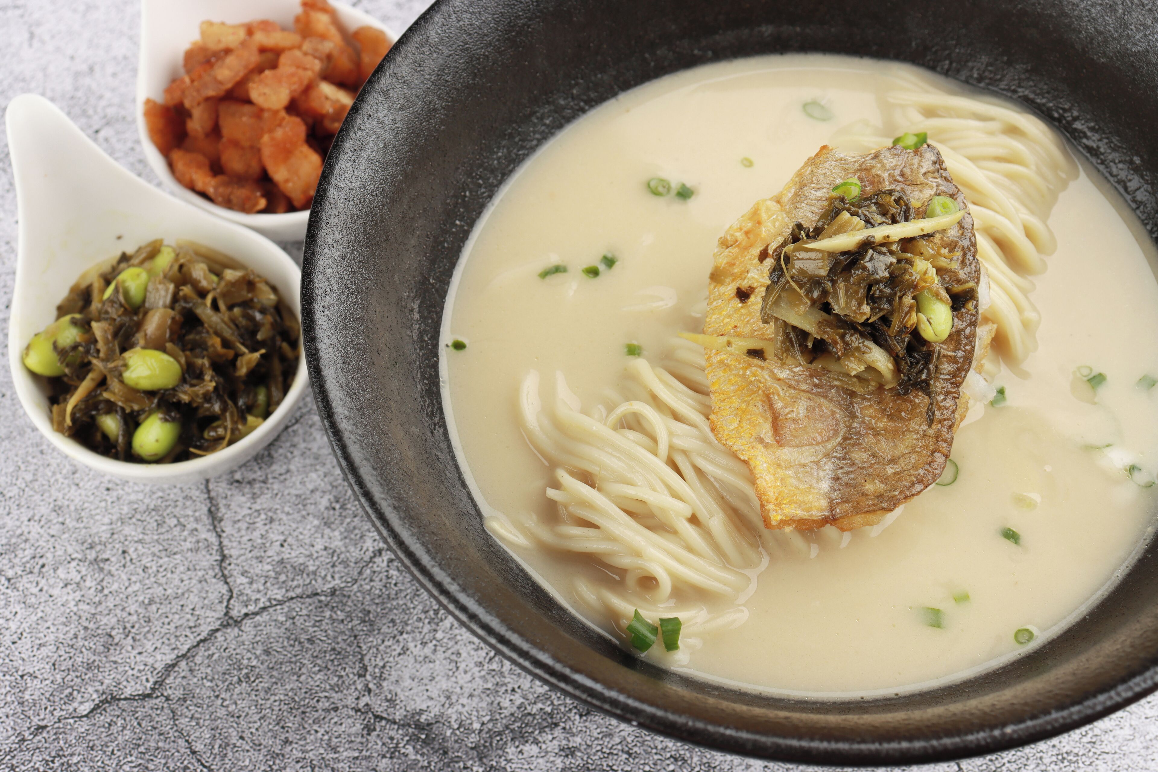 Uma tigela de sopa com macarrão e um pedaço de peixe, acompanhada por duas pequenas tigelas de acompanhamentos.