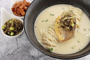 Uma tigela de sopa com macarrão e um pedaço de peixe, acompanhada por duas pequenas tigelas de acompanhamentos.