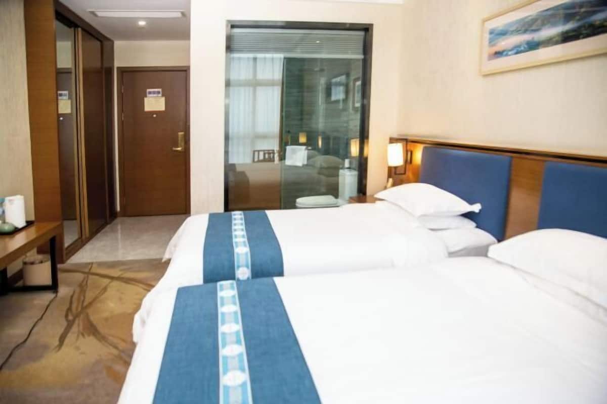 Quarto de hotel com duas camas, uma escrivaninha, uma cadeira, uma mesinha de cabeceira, um espelho e uma porta.