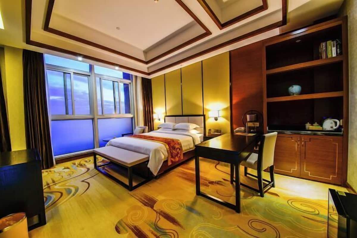 Quarto de hotel com cama, mesa de cabeceira, cadeira e vista do céu através de grandes janelas.