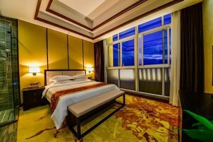 Quarto de hotel com uma cama grande, luminárias de cabeceira, um banco, box de vidro para o chuveiro e vista do céu noturno através de uma janela ampla.
