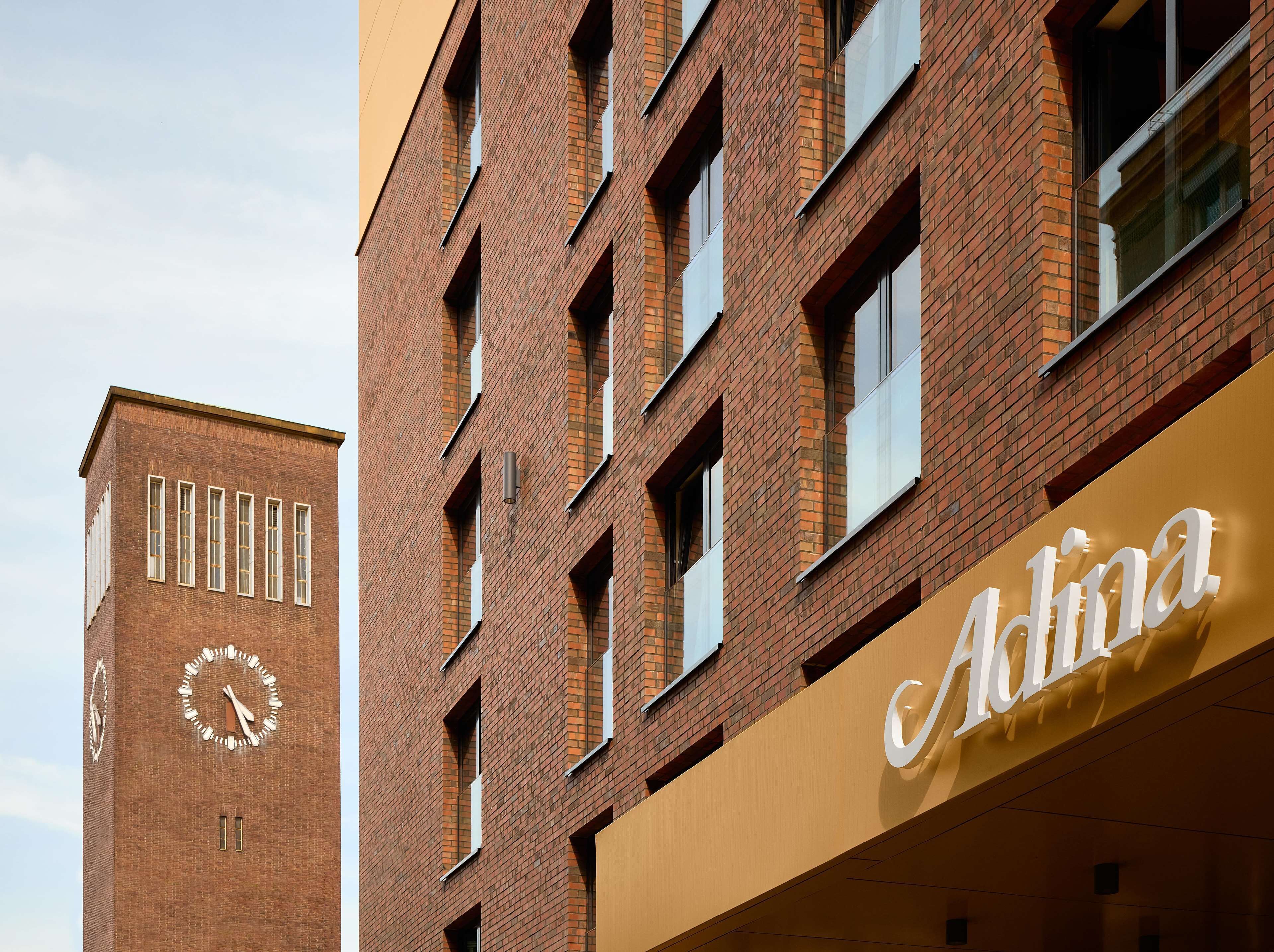 Edifício de tijolos com um relógio no lado esquerdo e uma placa com o nome 'Adina' no lado direito.
