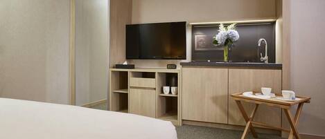 Quarto de hotel compacto com cama, televisão, pequena cozinha com pia e mesa com xícaras.