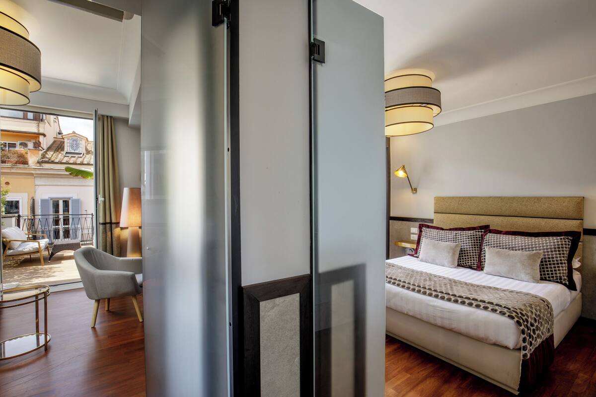 Quarto de hotel moderno com uma cama grande, área de estar com uma poltrona e varanda com vista para edifícios.