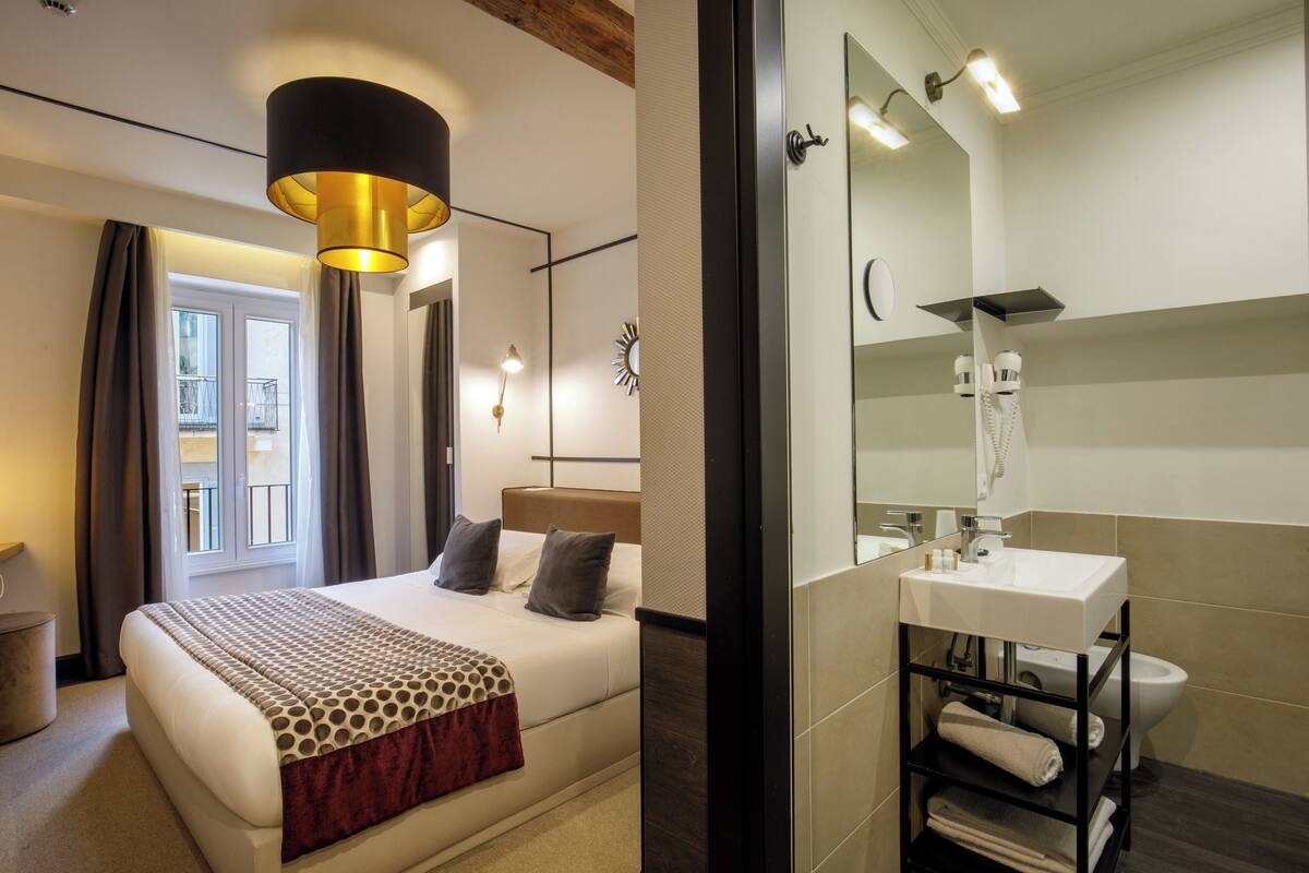 Quarto de hotel moderno com uma cama grande, banheiro com pia e vaso sanitário, e varanda com cortinas.
