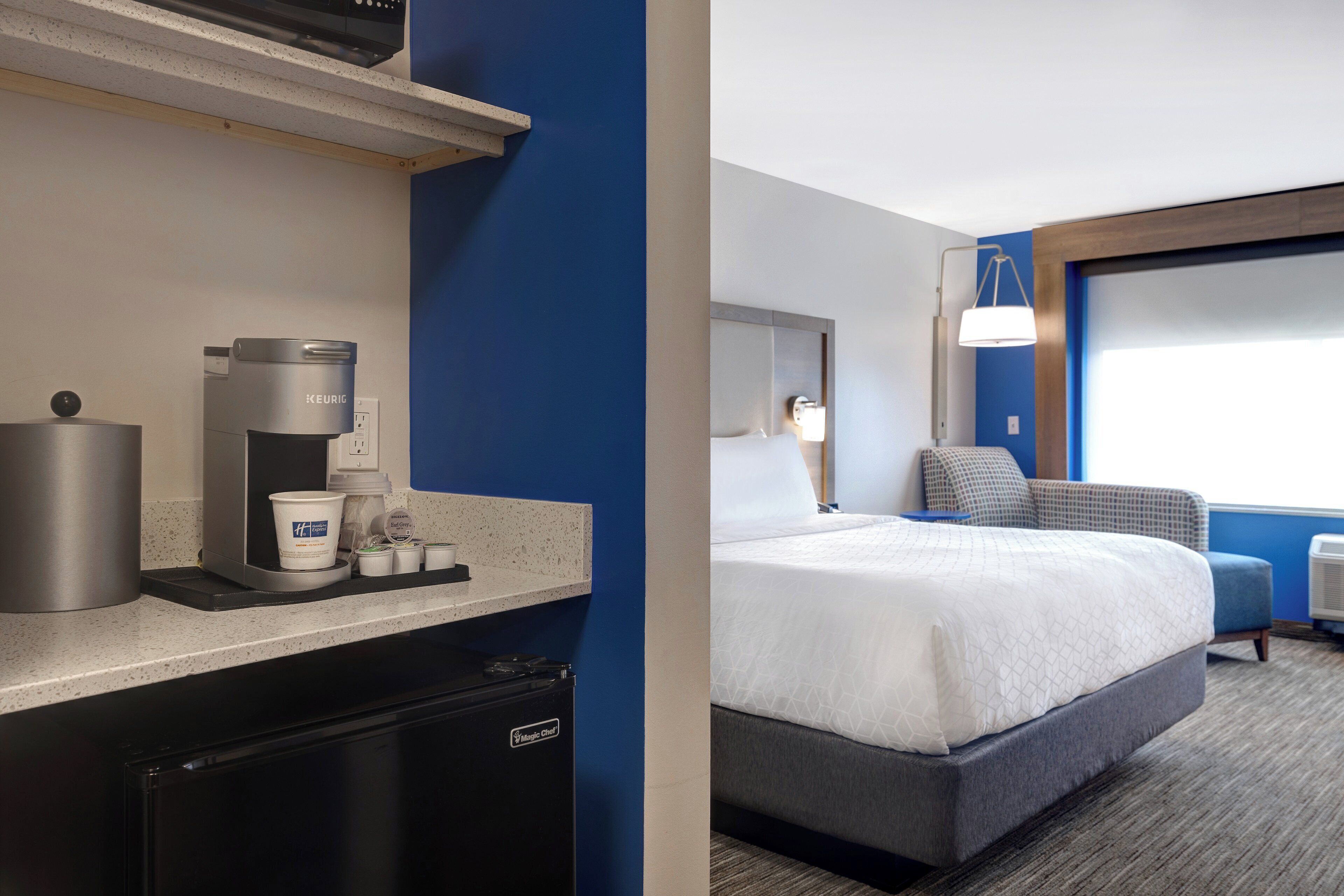 Quarto de hotel com cozinha compacta, cama, mesa de cabeceira e janela.