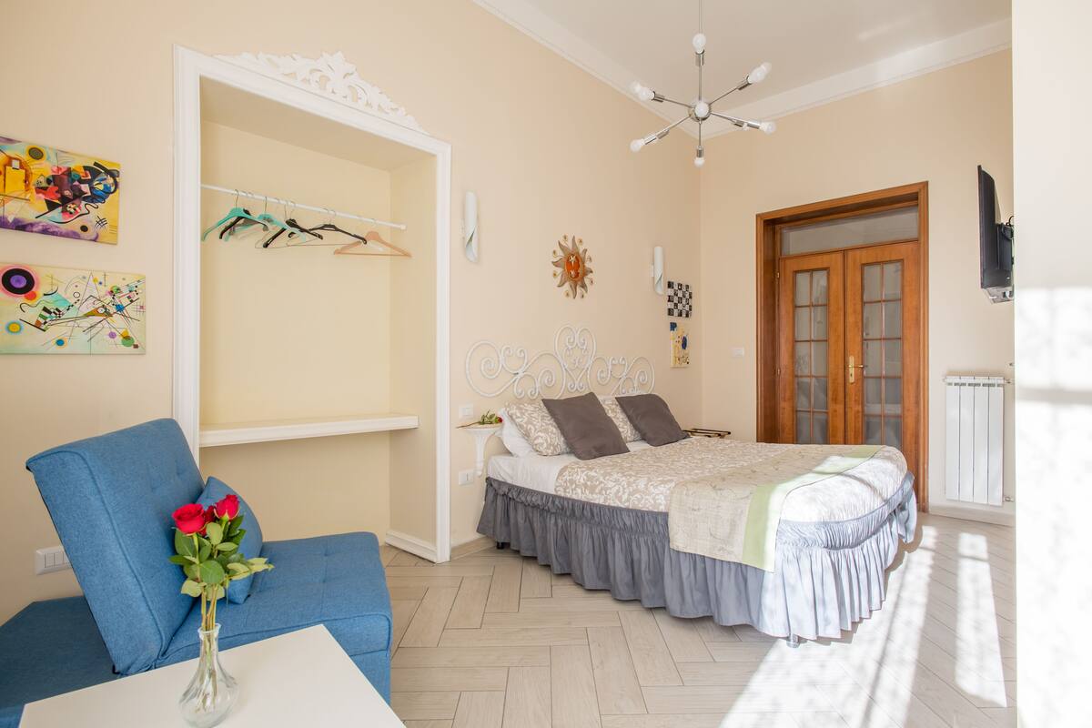 Quarto com cama, cadeira azul, mesa branca com um vaso de flores e obras de arte na parede.